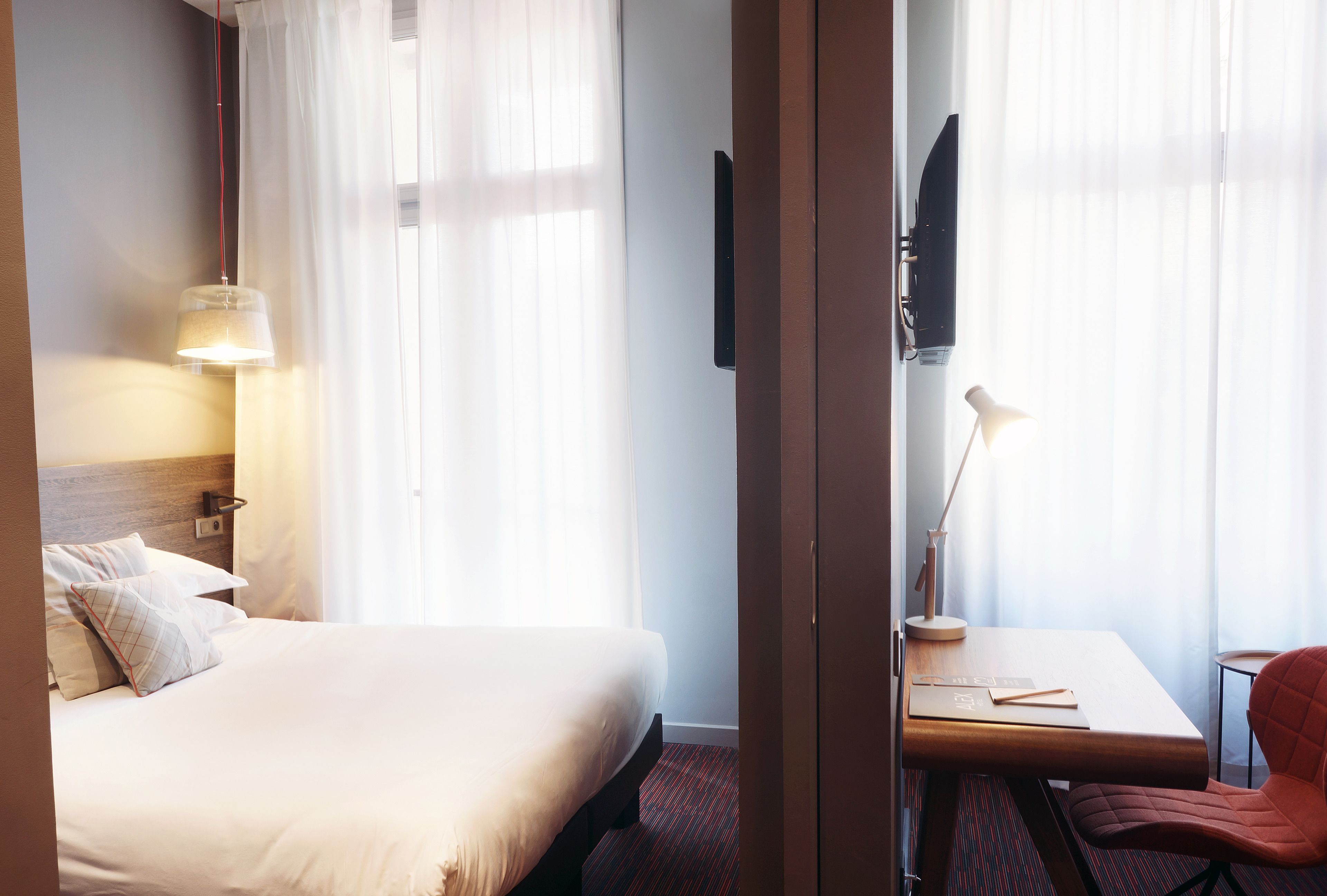 Quarto de hotel com cama, escrivaninha, cadeira, luminária e televisão fixada na parede.