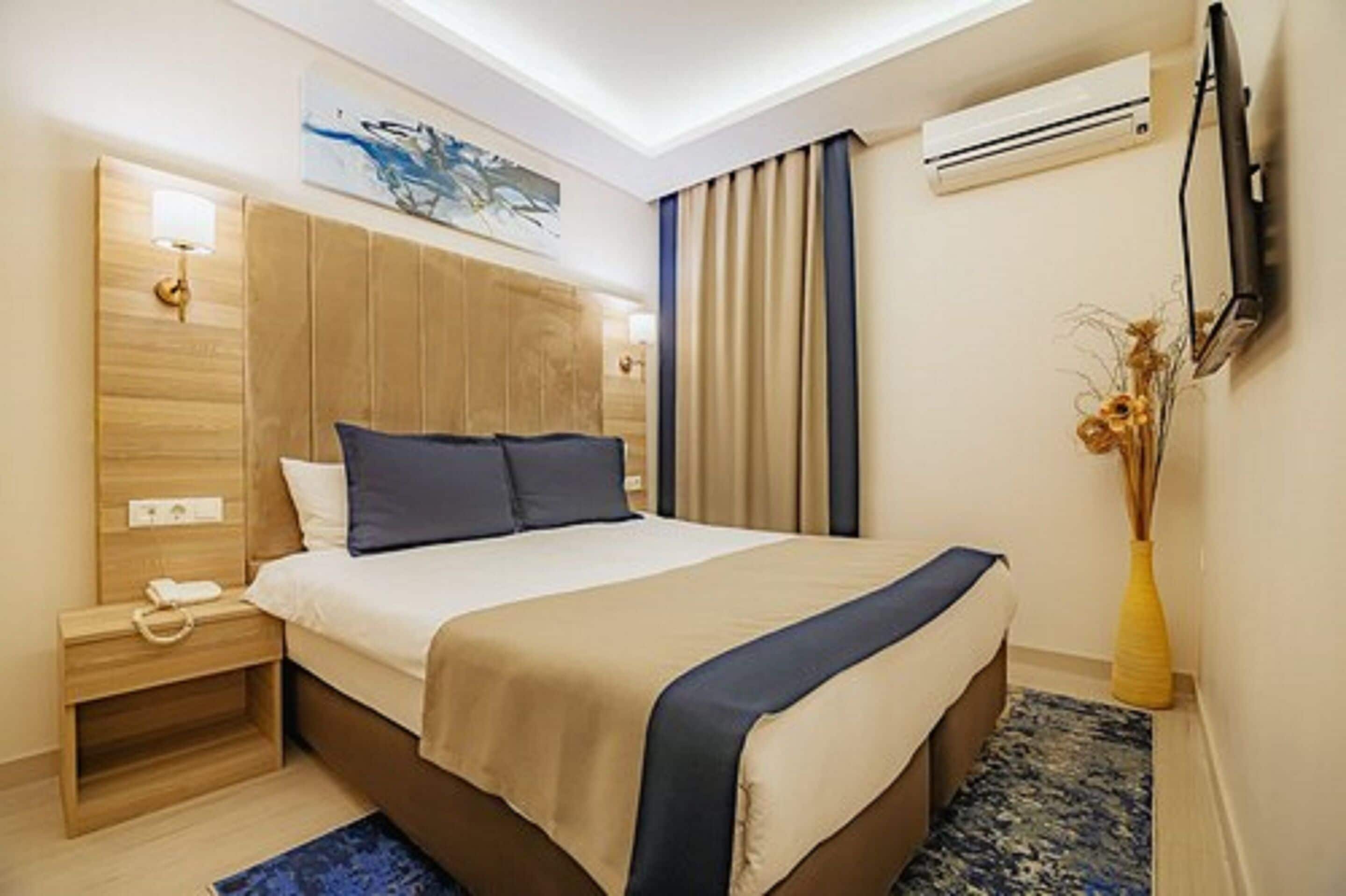 Quarto de hotel com cama, mesa de cabeceira, luminária, aparelho de ar condicionado e televisão fixada na parede.