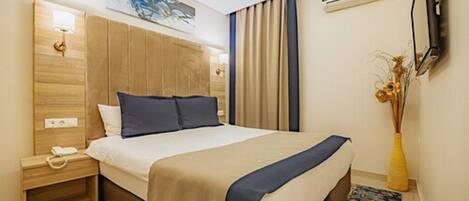 Quarto de hotel com cama, mesa de cabeceira, luminária, aparelho de ar condicionado e televisão fixada na parede.