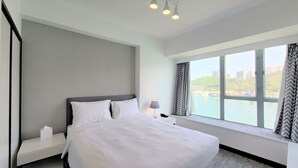 Quarto de hotel moderno com uma cama grande, uma mesinha de cabeceira com abajur e vista para a cidade através de uma janela ampla.