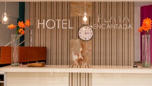 Uma área de recepção de hotel com design moderno, destacando-se um balcão de atendimento, um relógio e flores laranjas decorativas.