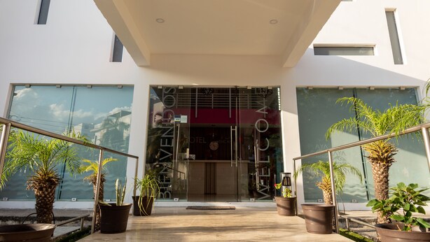 Uma entrada de hotel moderna com fachada de vidro, palmeiras e uma mesa de recepção.