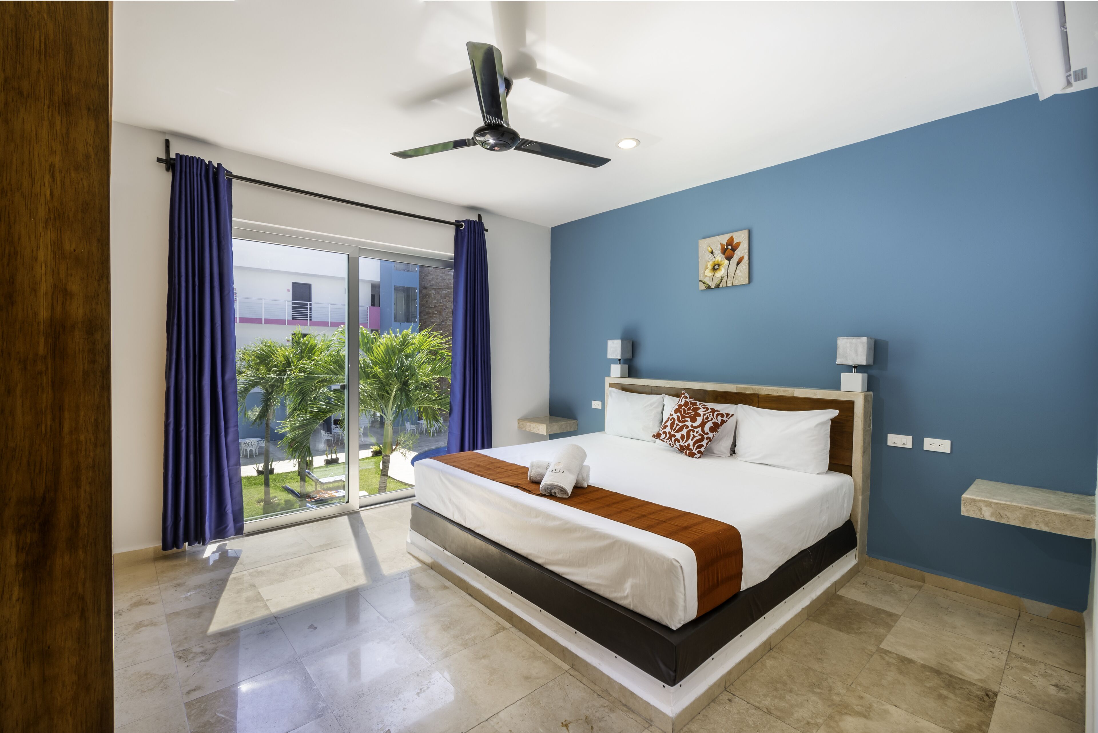 Quarto de hotel moderno com uma cama grande, ventoinha no teto, vista para o exterior e parede azul como detalhe.