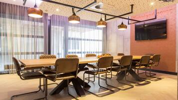 Uma sala de conferência com uma mesa comprida, várias cadeiras, iluminação pendente, uma televisão de tela plana e grandes janelas com cortinas finas.