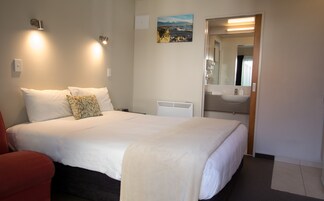 Quarto de hotel com uma cama, uma cadeira vermelha, um quadro e um banheiro visível através de uma porta aberta.