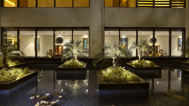 Vista noturna de um saguão de hotel com piscina refletora, plantas em vasos e fachada de um edifício grande com janelas e portas.