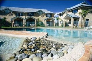 Piscina com borda rochosa e um edifício ao fundo com várias varandas.