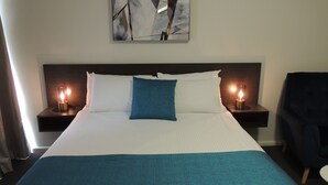 Quarto de hotel com uma cama grande, duas mesas de cabeceira com abajures, um cobertor azul e uma pintura na parede.