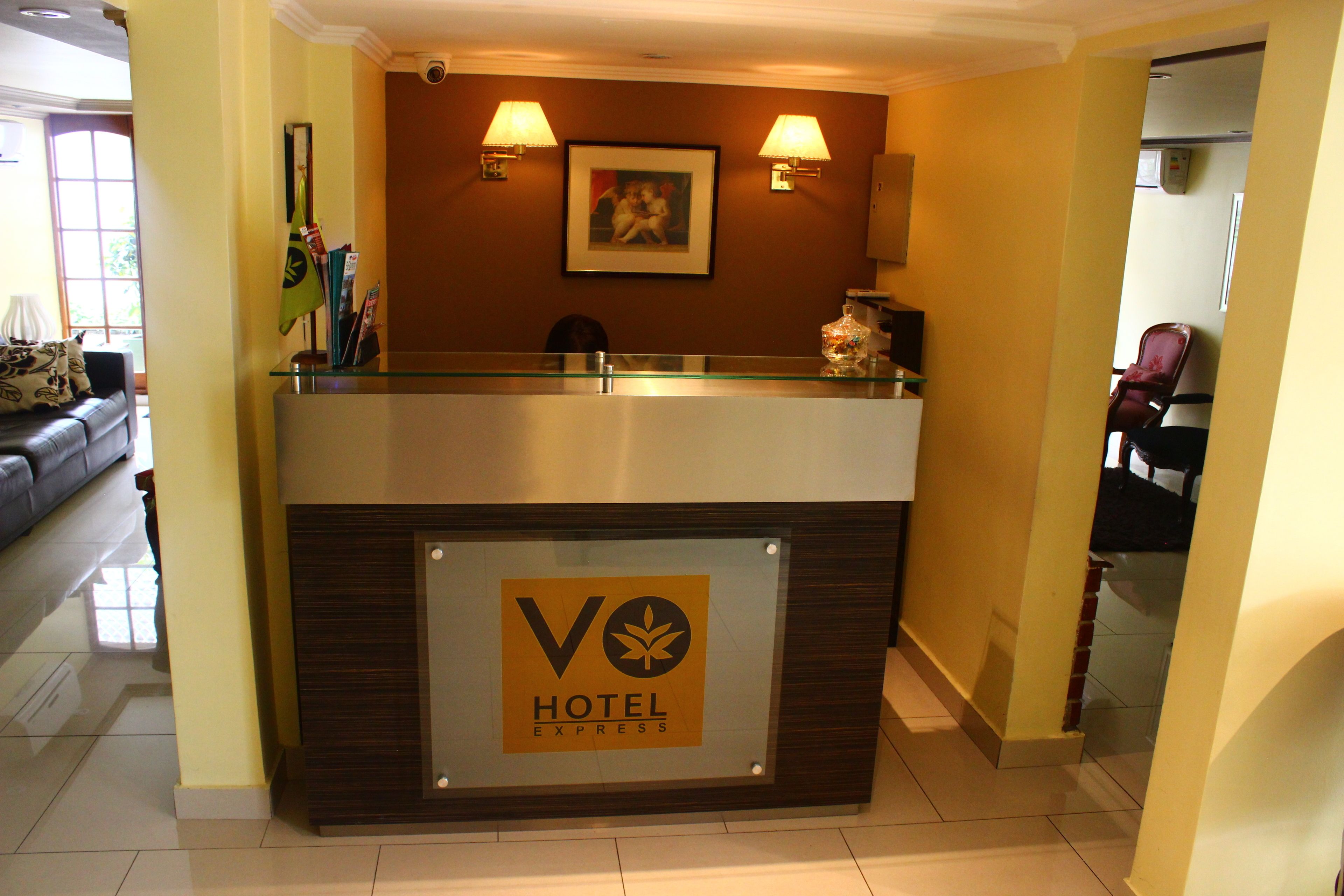 Bancada de recepção de hotel com tampo de vidro, uma moldura com imagem acima e duas luminárias fixadas na parede.