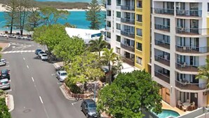 Um motel de vários andares com piscina, situado perto de uma praia com águas azul-claras e uma estrada com carros estacionados.