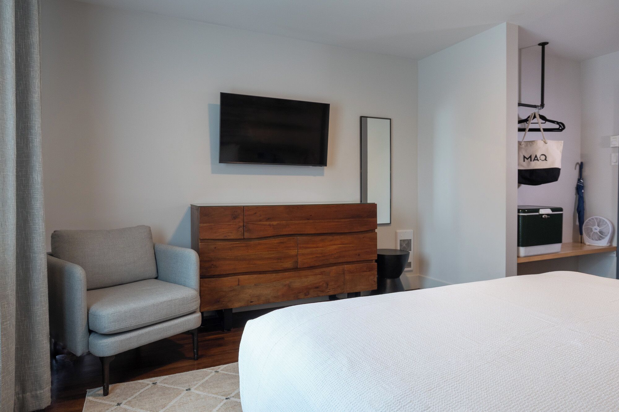 Quarto com cama, cabeceira de madeira, poltrona cinza, mesa pequena e televisão fixada na parede.