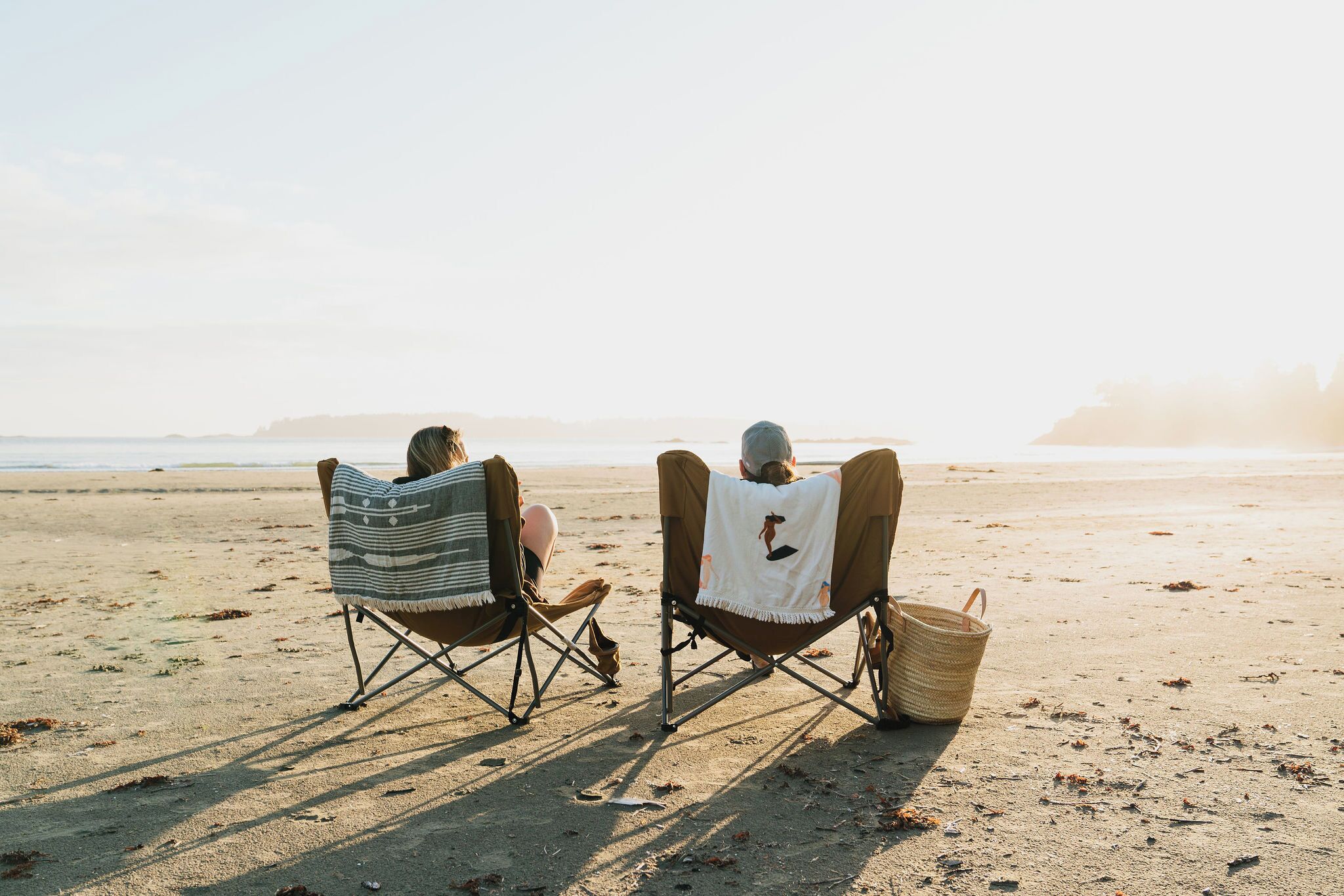 Duas pessoas sentadas em cadeiras de praia, olhando o oceano, com uma praia e paisagens distantes ao fundo.