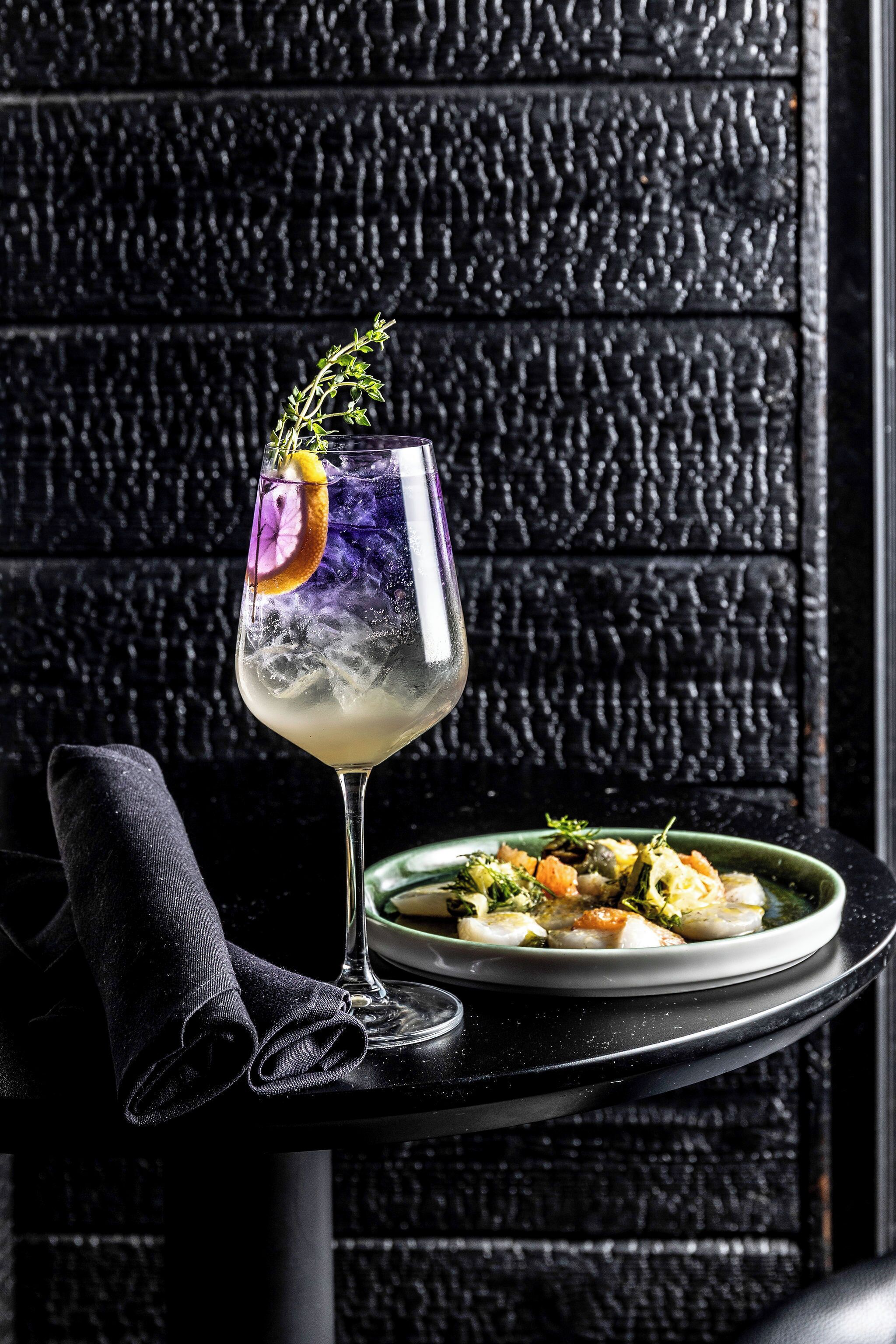 Um copo de cocktail com uma guarnição sobre uma mesa com um prato de comida.