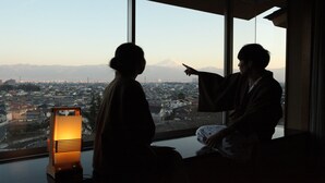 Duas pessoas sentadas perto de uma grande janela com vista para a cidade e uma montanha ao longe.