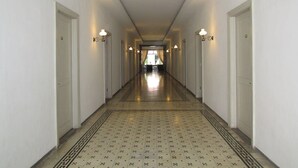 Um corredor comprido e estreito, com portas em ambos os lados, luminárias nas paredes e um chão estampado.