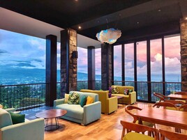 Uma varanda espaçosa com vista para uma paisagem urbana e montanhas, equipada com sofás, cadeiras e uma mesa.