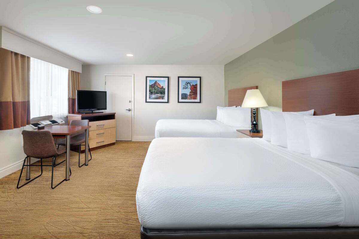 Quarto de hotel com duas camas, uma escrivaninha com cadeira, uma televisão e duas quadros na parede.