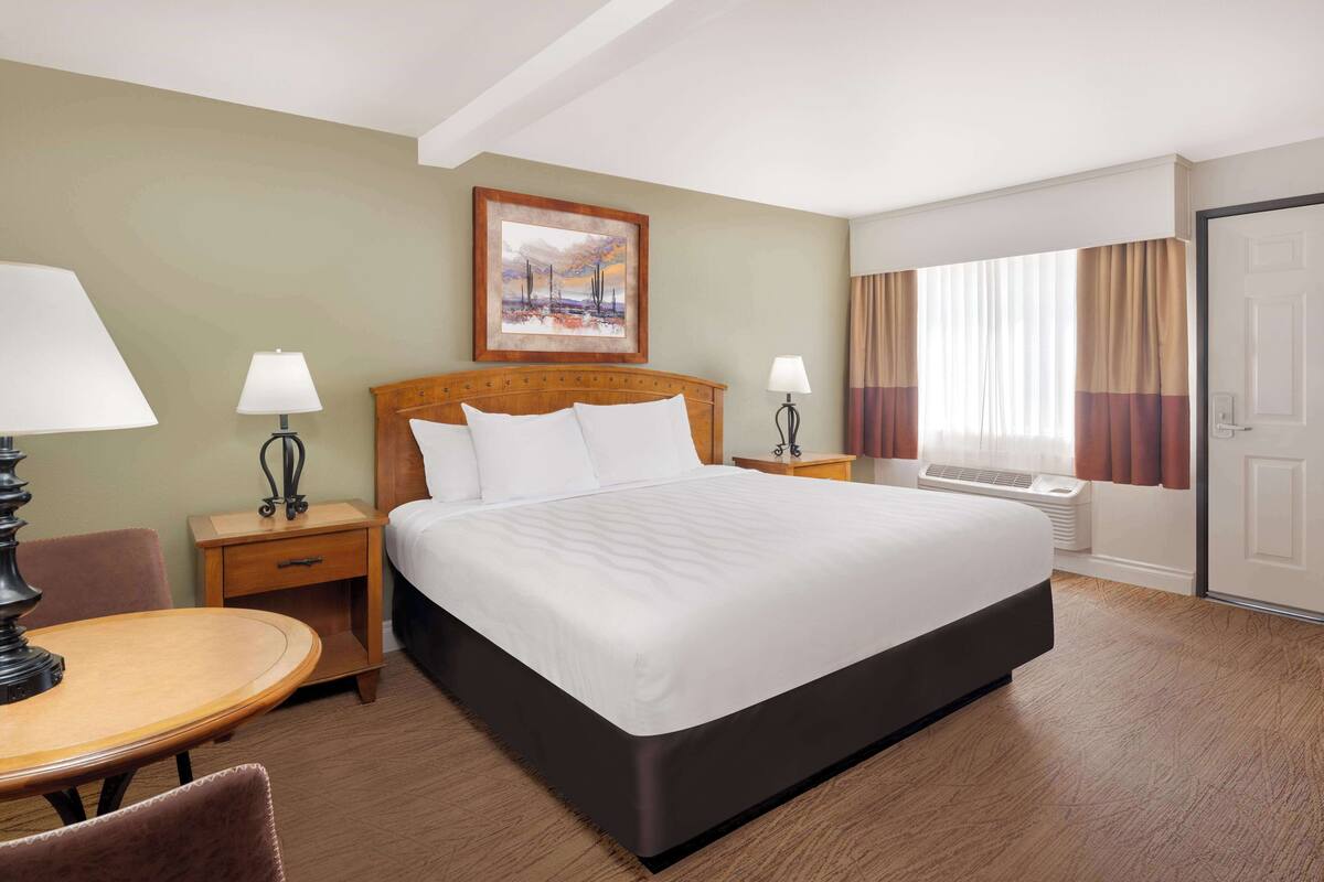 Quarto de hotel com uma cama grande, duas mesas de cabeceira com abajures, uma mesinha redonda com uma cadeira, um quadro na parede e uma janela com cortinas.