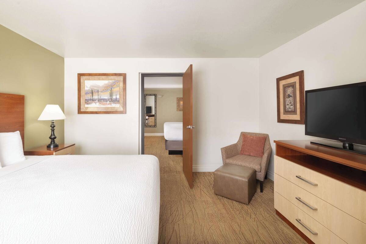 Quarto de hotel com cama, televisão, escrivaninha, cadeira e porta que dá para outro espaço.
