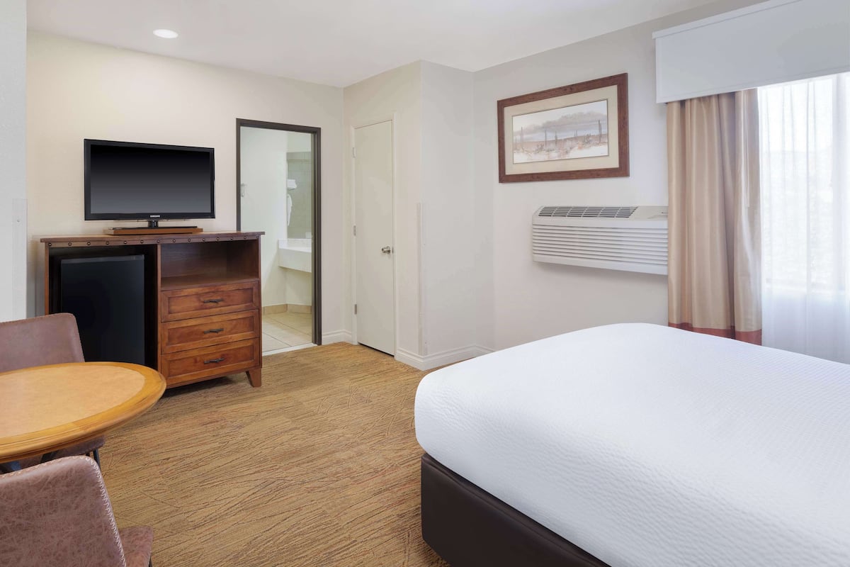 Quarto de hotel com cama, televisão em móvel, escrivaninha com cadeira, guarda-roupa e porta que dá para outro quarto.