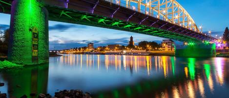 Uma ponte com estrutura metálica e luzes, atravessando um corpo d'água calmo, com um horizonte urbano ao fundo.