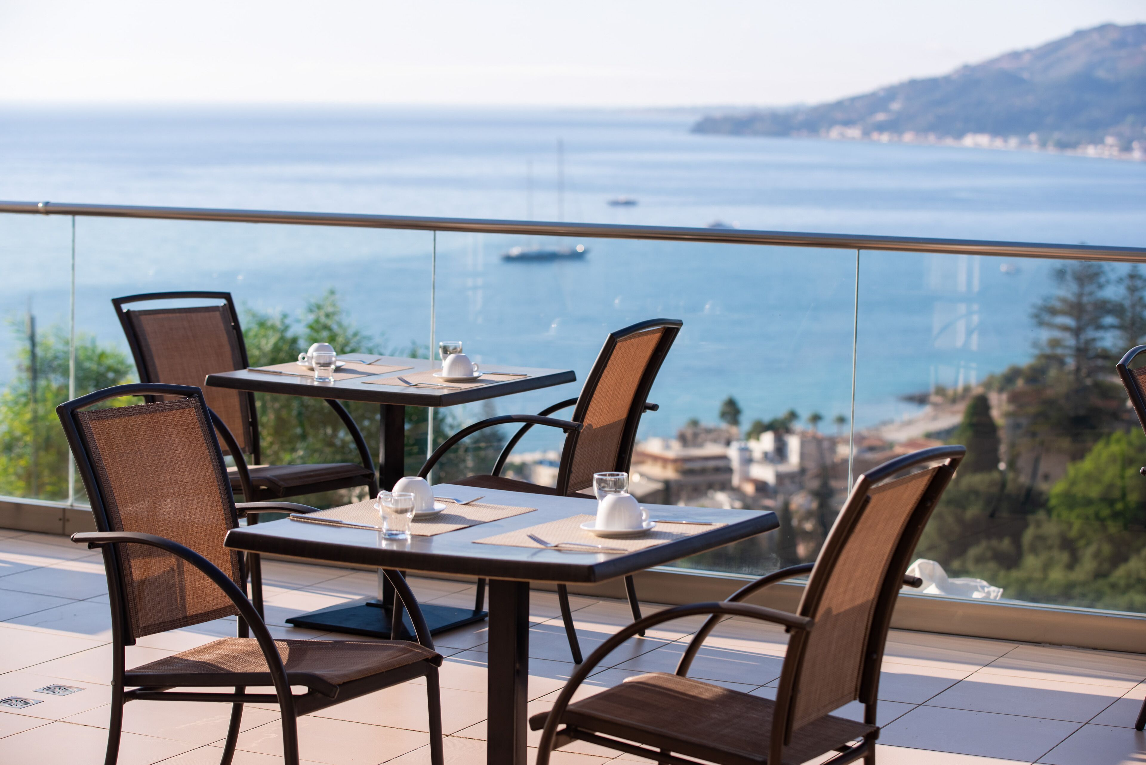 Terraço com mesas e cadeiras, vista para a costa com barcos e edifícios.