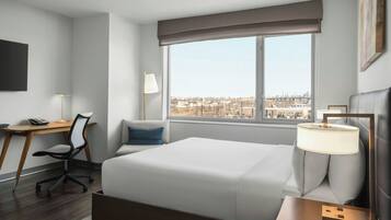Quarto de hotel com cama, escrivaninha, cadeira, luminária e vista da cidade.