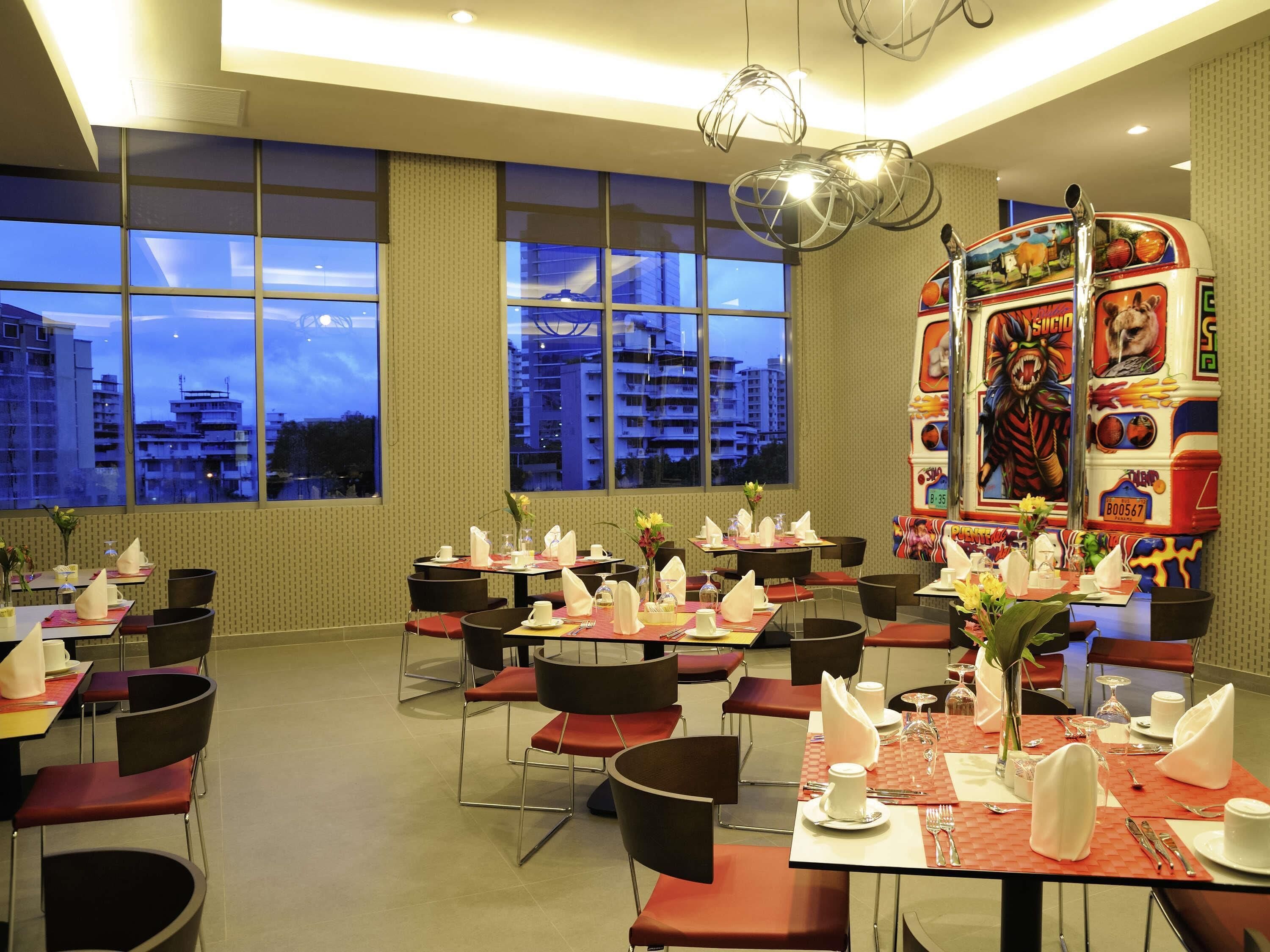 Um restaurante com mesas preparadas para refeições, grandes janelas com vista para a cidade e um mural colorido na parede.