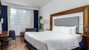 Quarto de hotel com uma cama grande, uma escrivaninha com cadeira, um sofá, uma televisão e uma janela com cortinas.