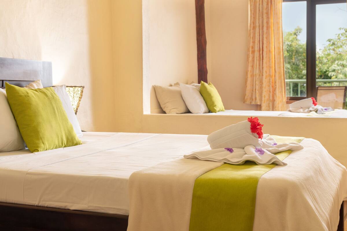 Quarto de hotel com cama, almofadas, janela com cortinas e vista para o verde lá fora.