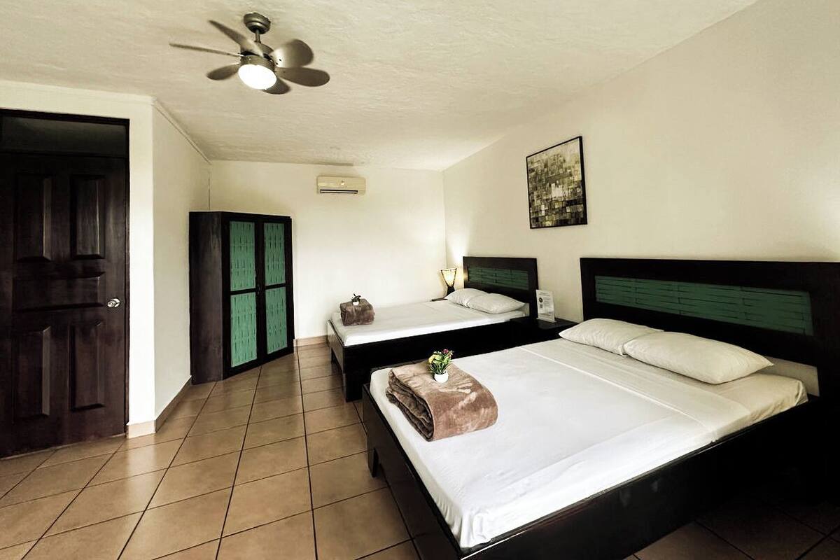 Quarto de hotel com duas camas de solteiro, ventilador de teto, ar condicionado fixado na parede e porta com painel verde.