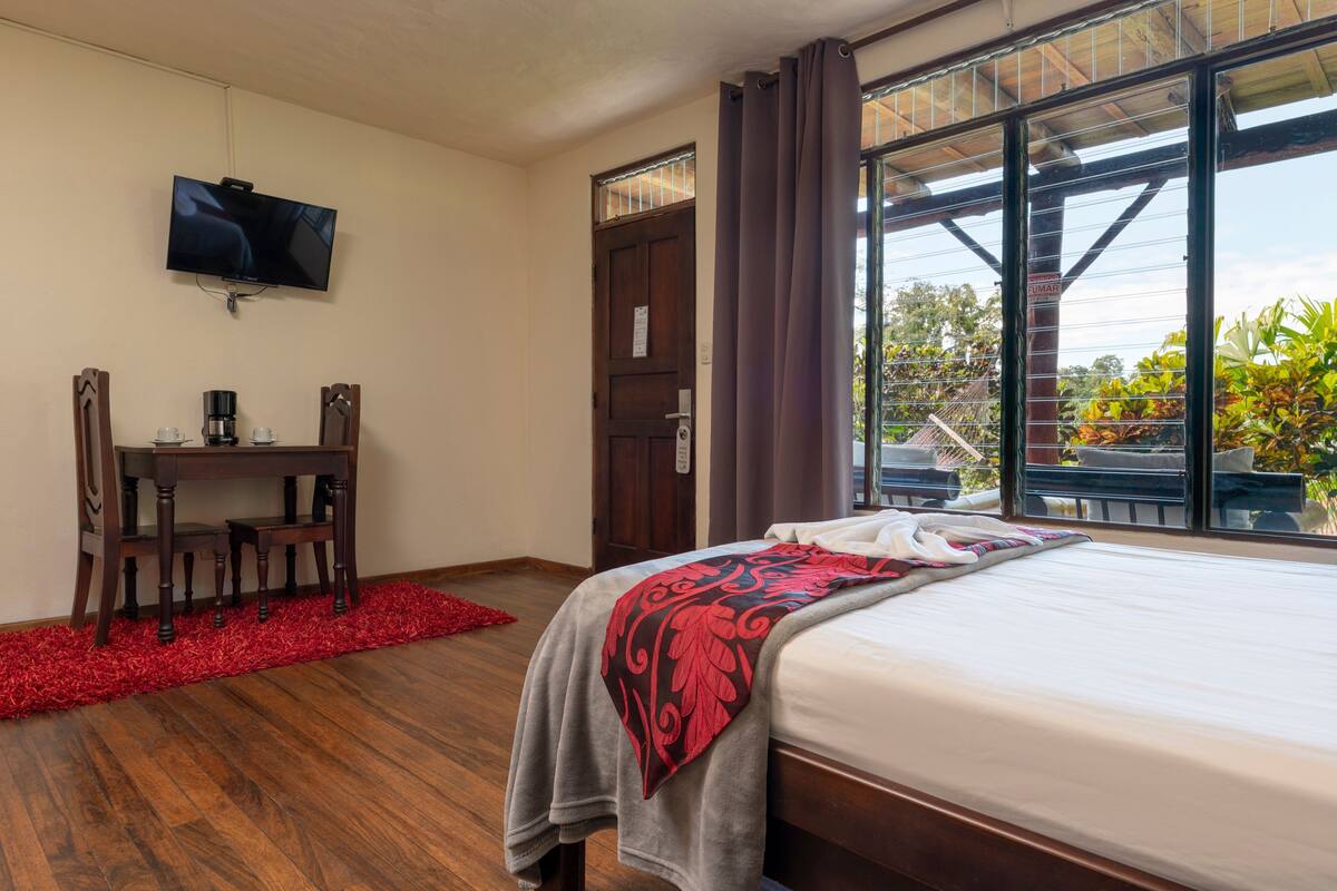 Quarto de hotel com cama, escrivaninha com cafeteira, TV na parede e uma grande janela.