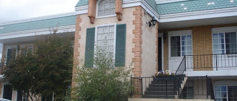 Um edifício de dois andares com telhado verde, fachada de tijolos e uma pequena varanda com guarda-corrimão preto.