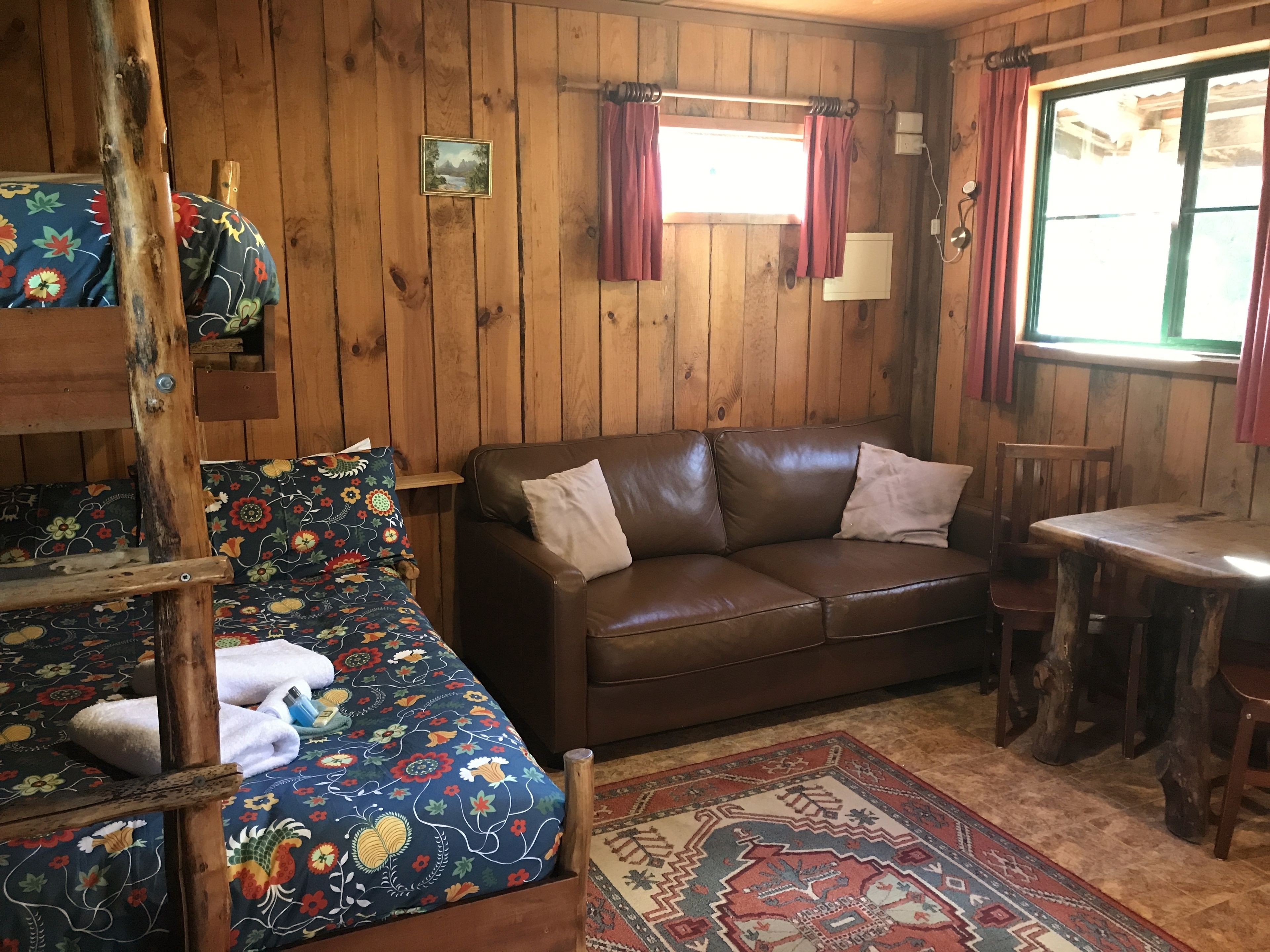 Interior de uma cabana de madeira com um sofá de couro marrom, uma cama com estampa floral, uma mesa de jantar rústica e uma cadeira de madeira.