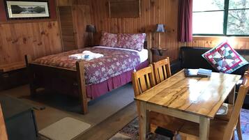 Quarto com painéis de madeira, uma cama, uma mesa e uma cadeira. Há uma janela com cortinas, um abajur e um quadro na parede.