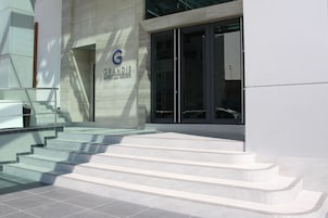 Entrada moderna de um hotel com fachada de vidro, escadaria visível e placa com a inscrição 'GRANDIS HOTEL'.