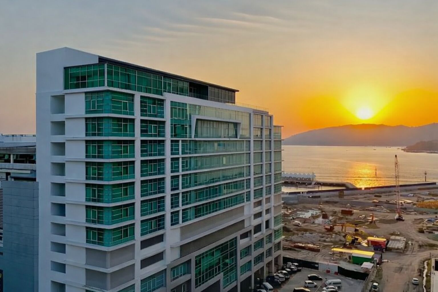 Um edifício moderno com vários andares e grandes janelas, com vista para um corpo de água e um pôr do sol.