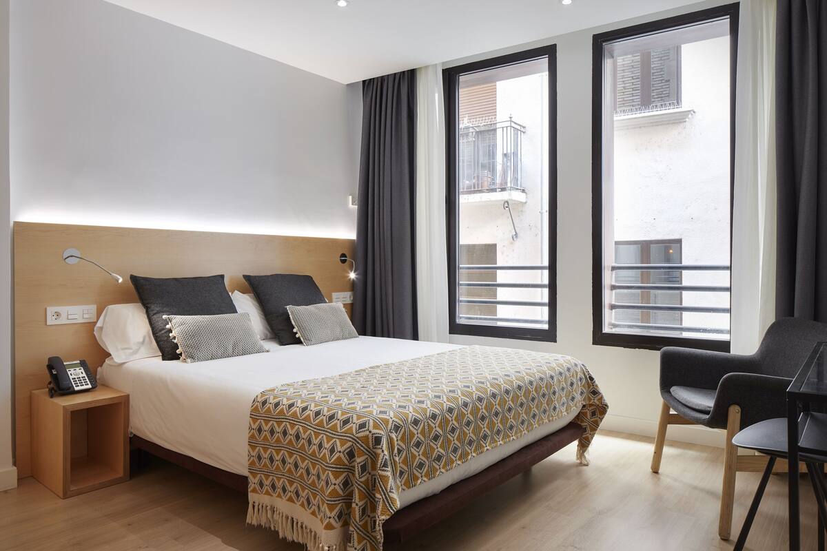 Quarto de hotel com cama, mesa de cabeceira com telefone, uma cadeira e janelas amplas com vista para uma varanda.