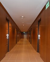 Corredor longo e estreito de hotel com portas de madeira em ambos os lados, piso ladrilhado e teto com iluminação embutida.