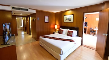 Quarto de hotel com uma cama grande, uma mesa de cabeceira com abajur, uma TV e uma porta que dá para uma sala de jantar.