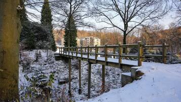 Uma ponte de madeira sobre uma área coberta de neve, com árvores e edifícios ao fundo.