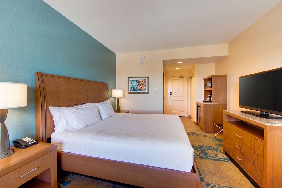 Quarto de hotel com cama, mesa de cabeceira, luminária, TV e uma pequena cozinha.