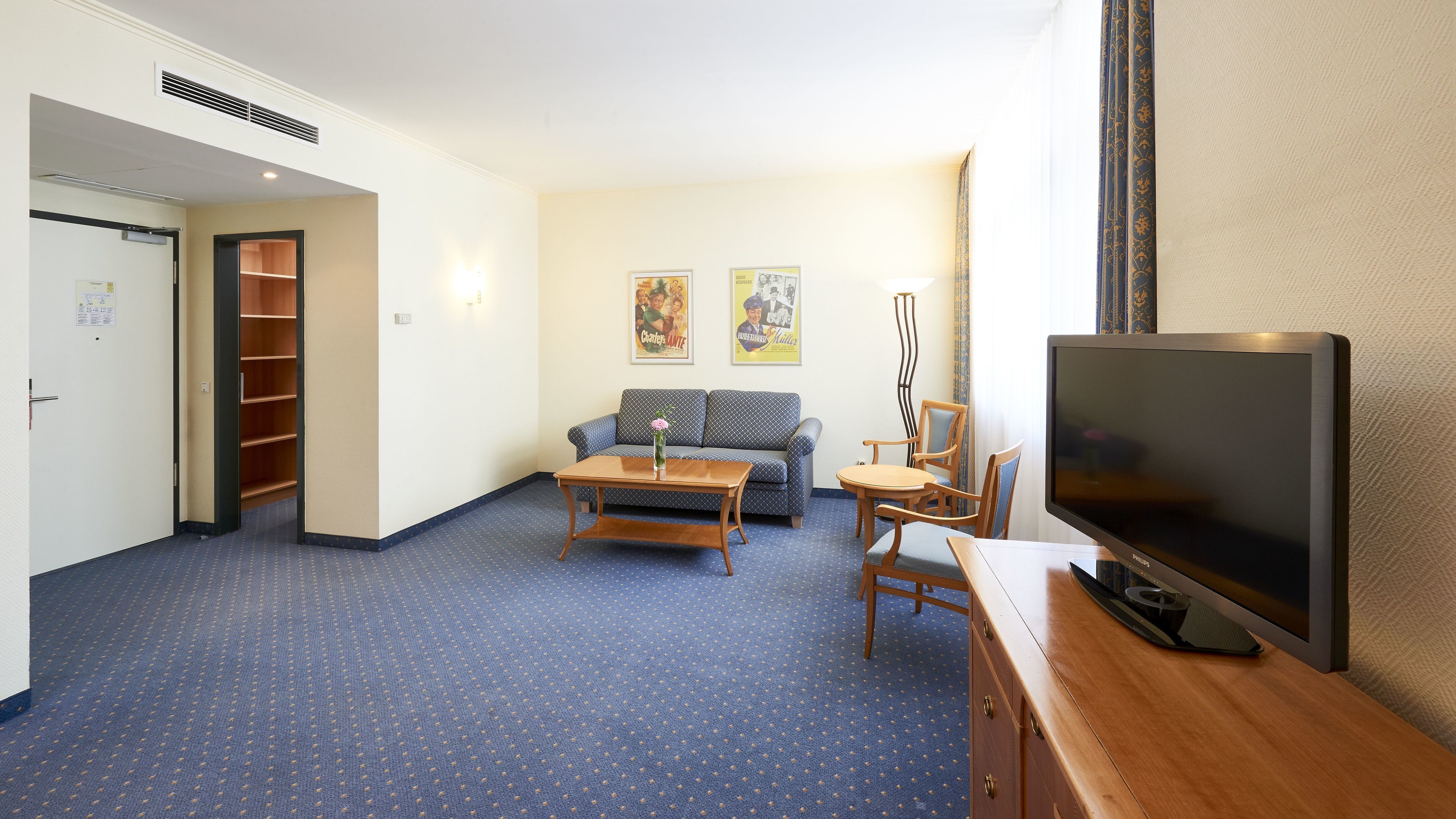 Quarto de hotel com sofá, mesa de centro, televisão, estante de livros e janela com cortinas.