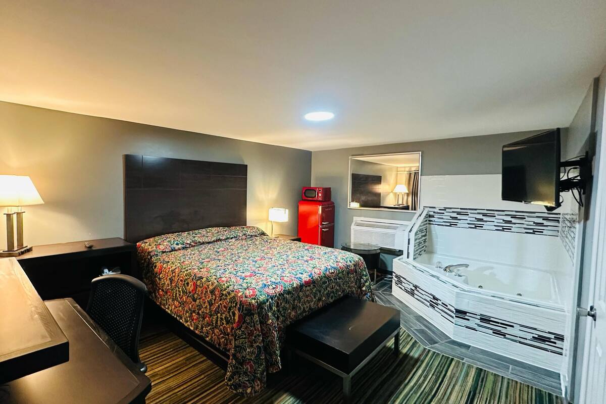 Quarto de hotel com cama, escrivaninha, cadeira, minibar vermelho, televisão, espelho e hidromassagem.