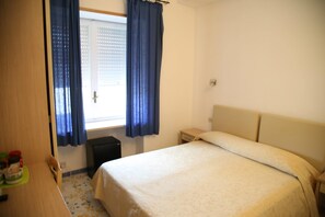 Quarto com cama, mesa de cabeceira, uma pequena mesa com cadeira e janela com cortinas.