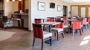 Espaço de refeições de hotel com várias mesas e cadeiras, um balcão de recepção e uma televisão na parede.