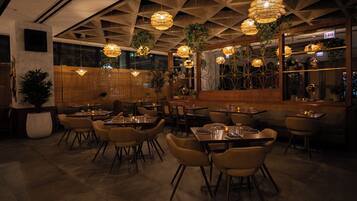 Interior de restaurante com várias mesas e cadeiras, luminárias pendentes e uma janela grande com vista.