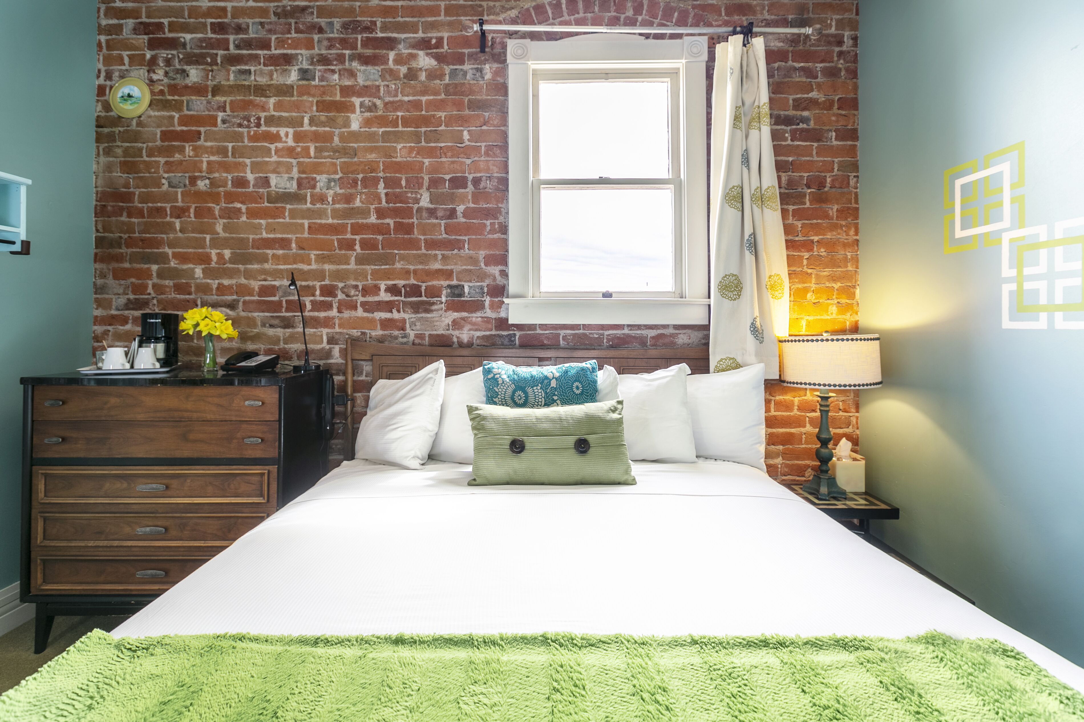 Quarto com parede de tijolos, janela com cortinas, cama com almofadas, cômoda e mesa de cabeceira com abajur.