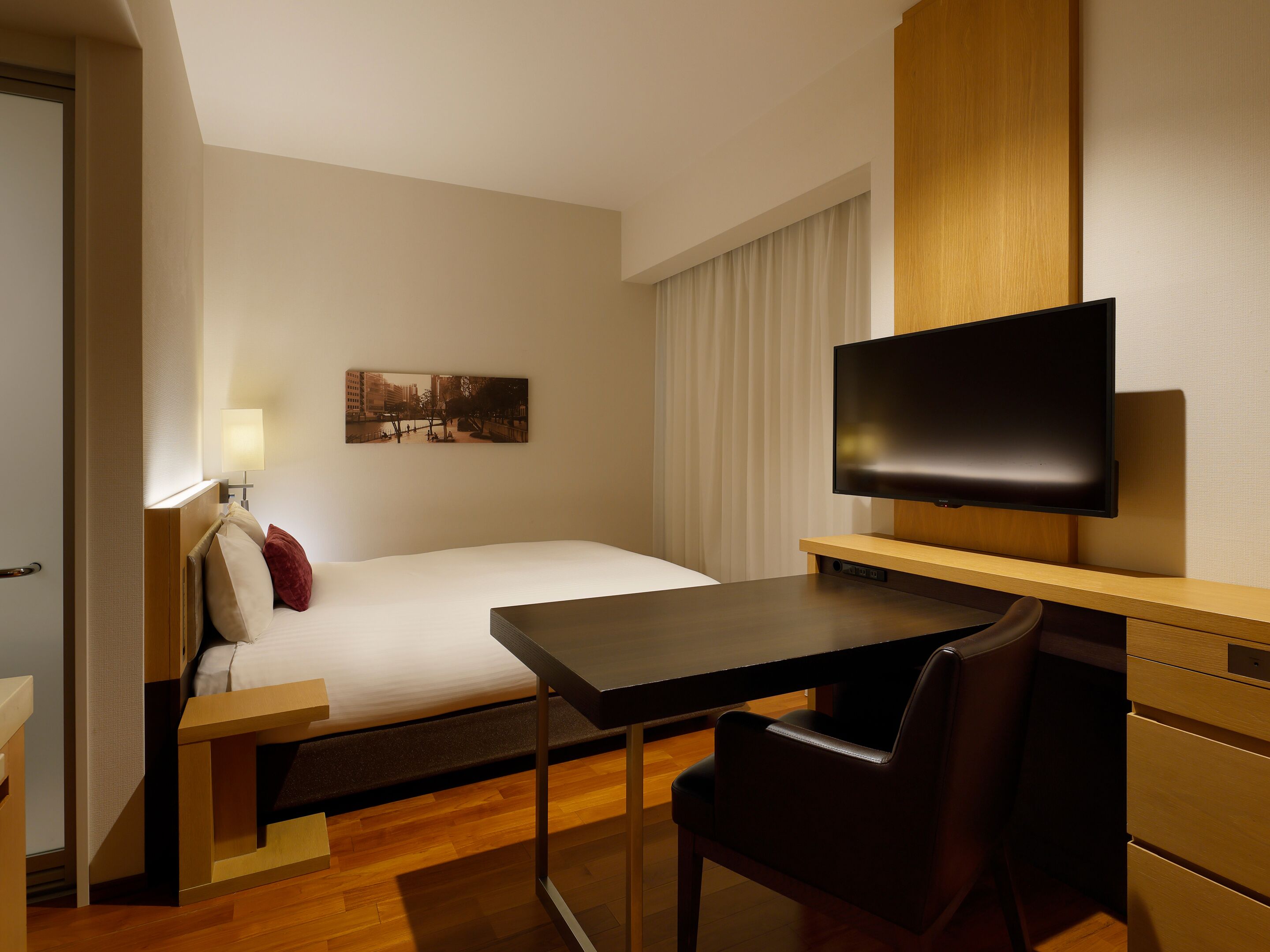 Quarto de hotel com cama, escrivaninha, cadeira, televisão e janela com cortinas.