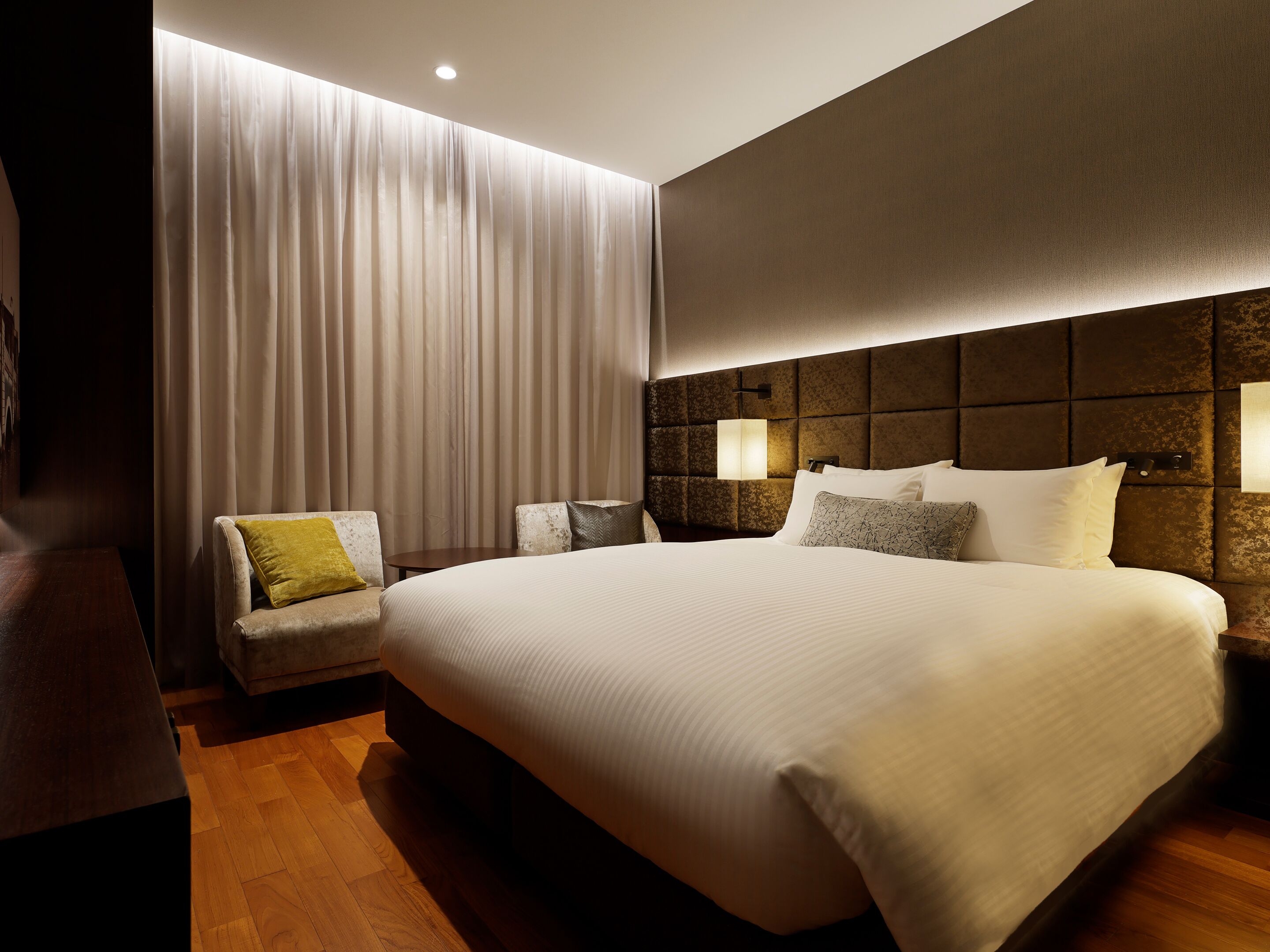 Quarto de hotel com uma cama grande, uma poltrona pequena, uma mesa de cabeceira e uma luminária na parede.